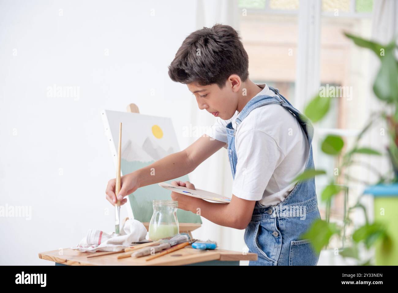 A young teenager is painting a picture of a landscape. He uses a brush ...