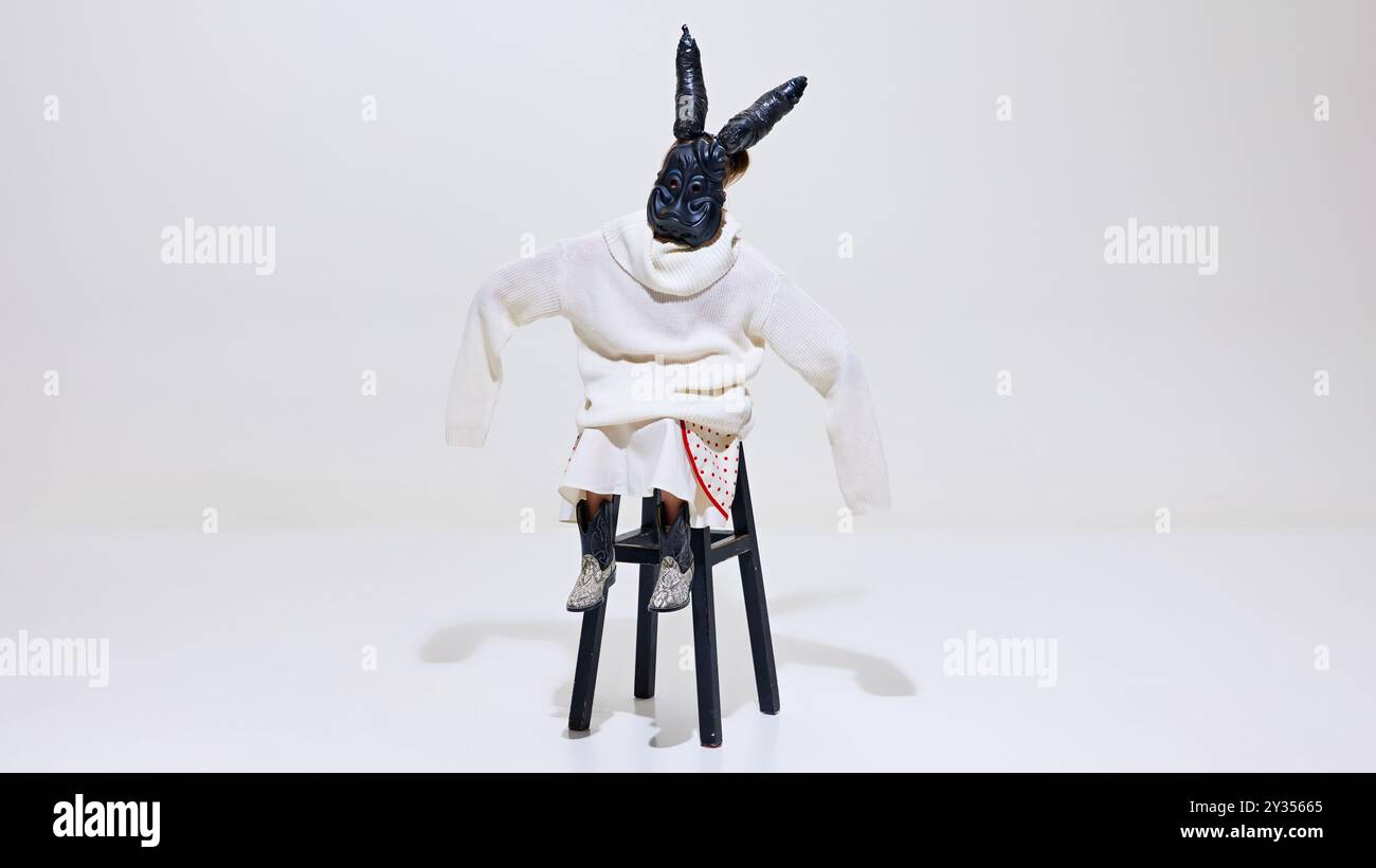 Figure with black, rabbit-like mask sitting on stool. Dressed in white ...