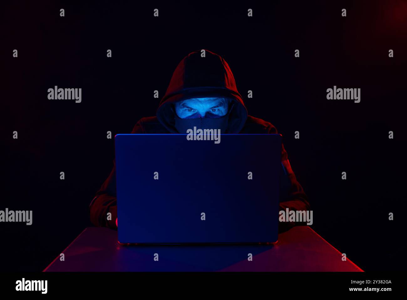 Cyber hacker in hood behind laptop on dark background with red blue ...
