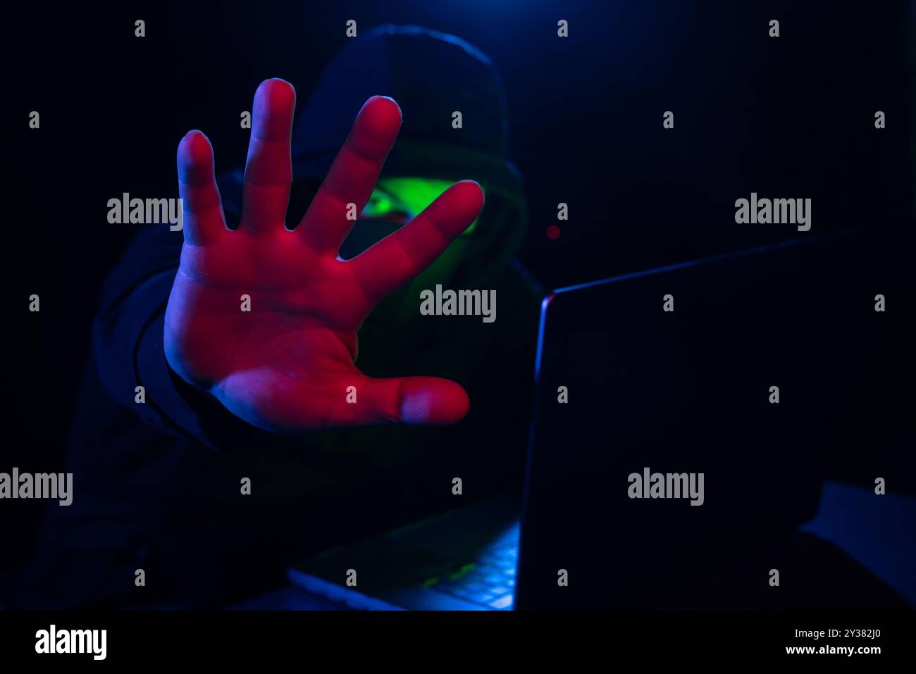 Cyber hacker in hood behind laptop puts hand forward hiding his face in ...