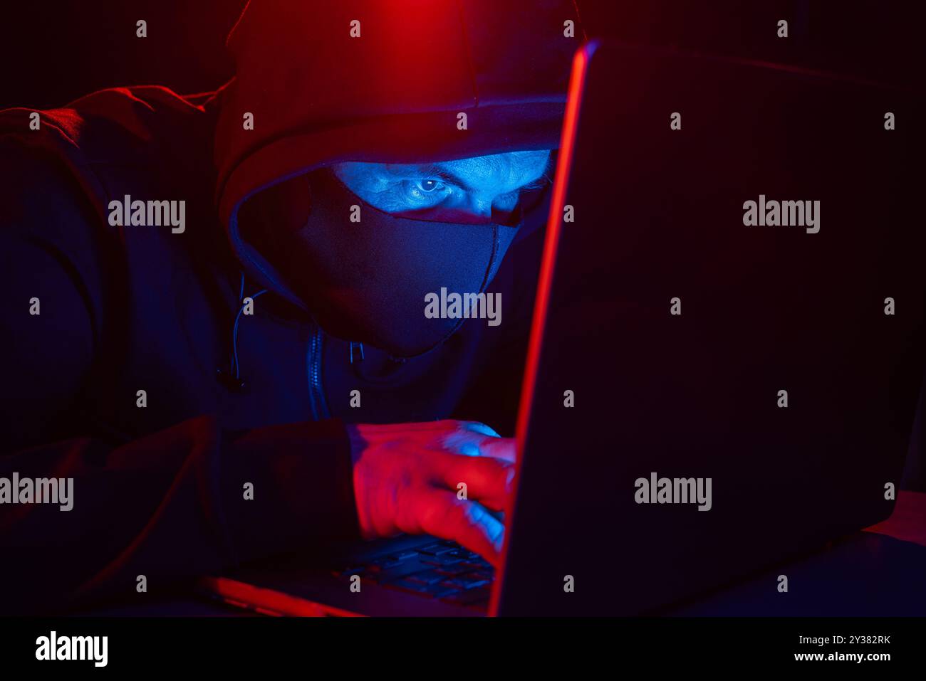 Cyber hacker in hood behind laptop on dark background with red blue light, anonymous spy ...