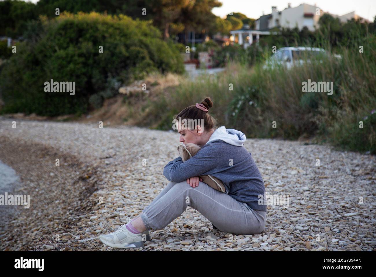 Young sad beautiful woman sitting on a pebble beach and hugs a pillow ...