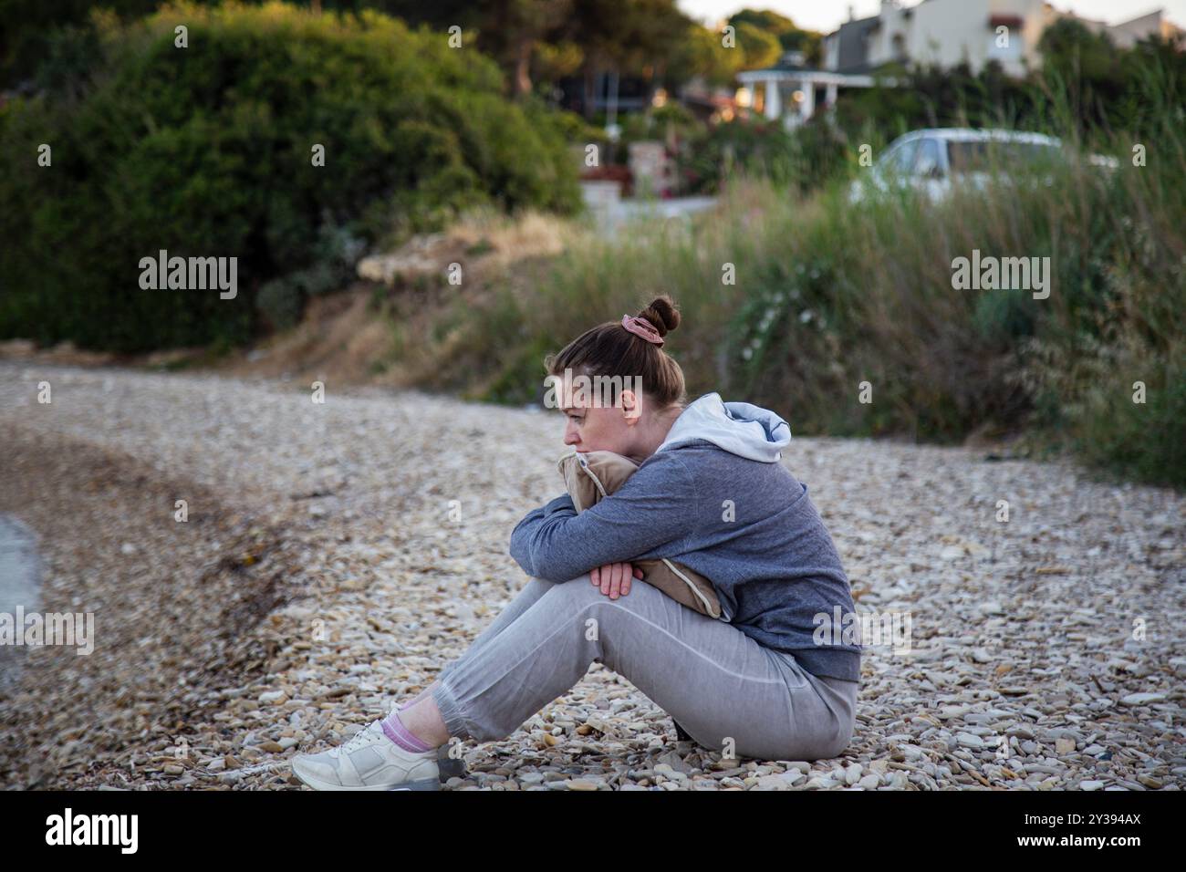 Young sad beautiful woman sitting on a pebble beach and hugs a pillow ...