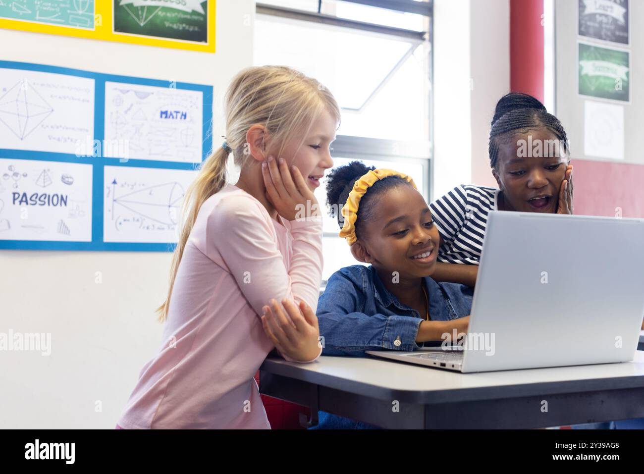 In school, three multiracial girls using laptop together in classroom, collaborating on project ...