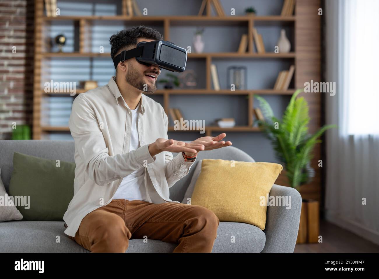 Young man sits on sofa wearing VR headset, experiencing virtual reality ...