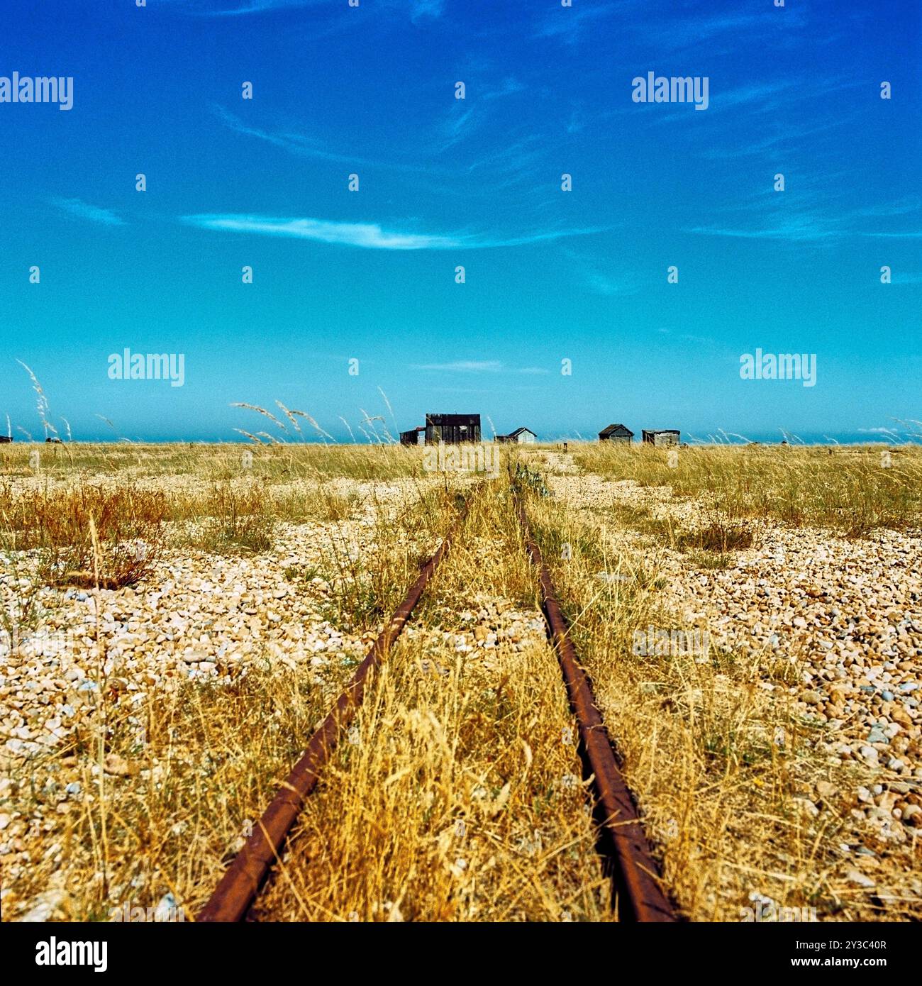 Abandoned railway tracks stretch towards rustic huts in an open field under a vibrant blue sky ...