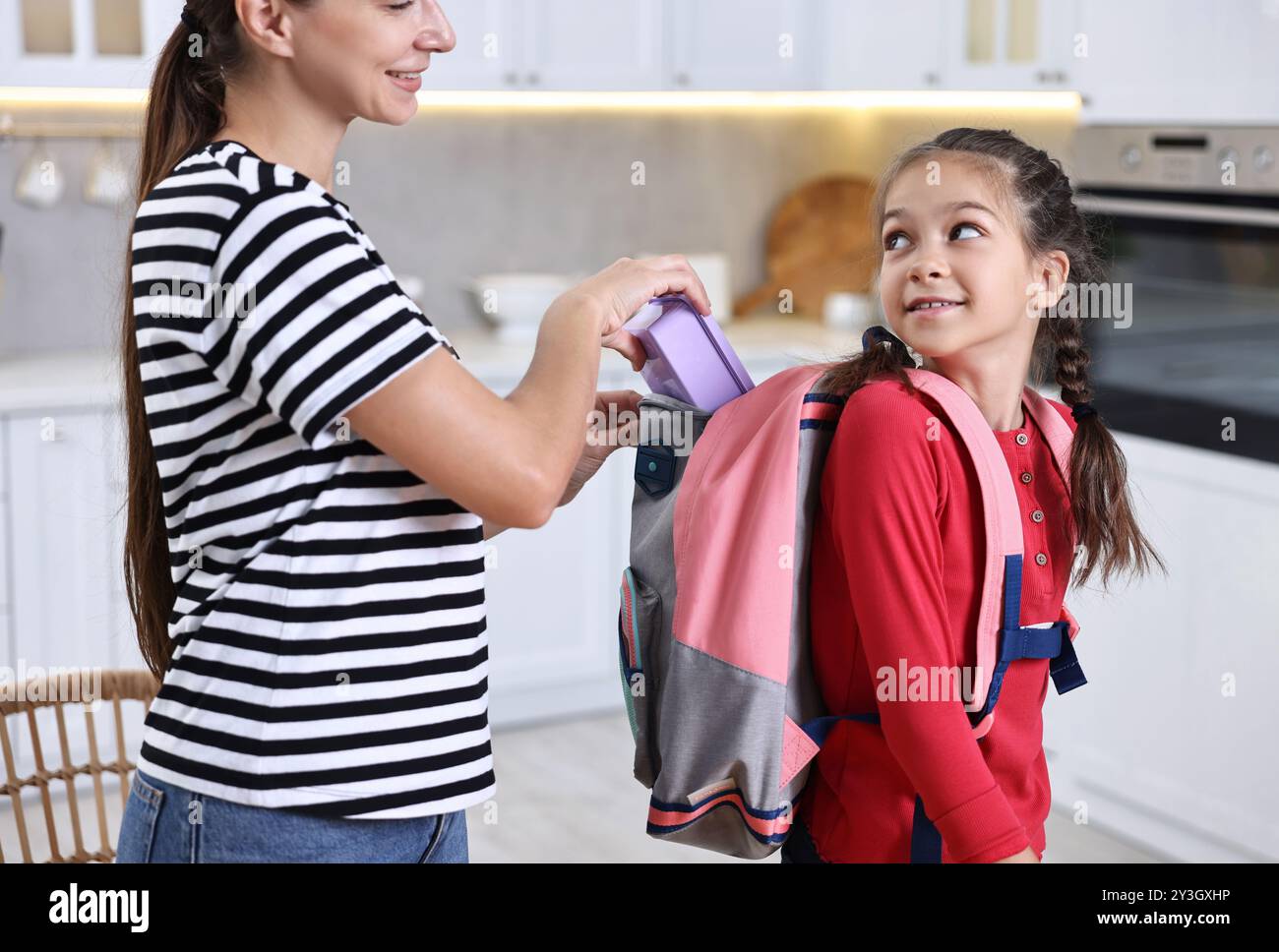 Mother putting lunch box into daughter`s backpack in kitchen. Preparing ...