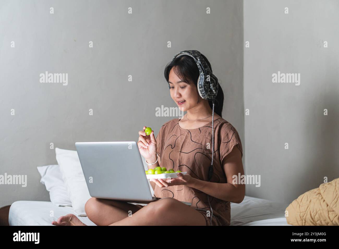 A young woman sitting on a bed, wearing headphones, using a laptop while enjoying green grapes ...