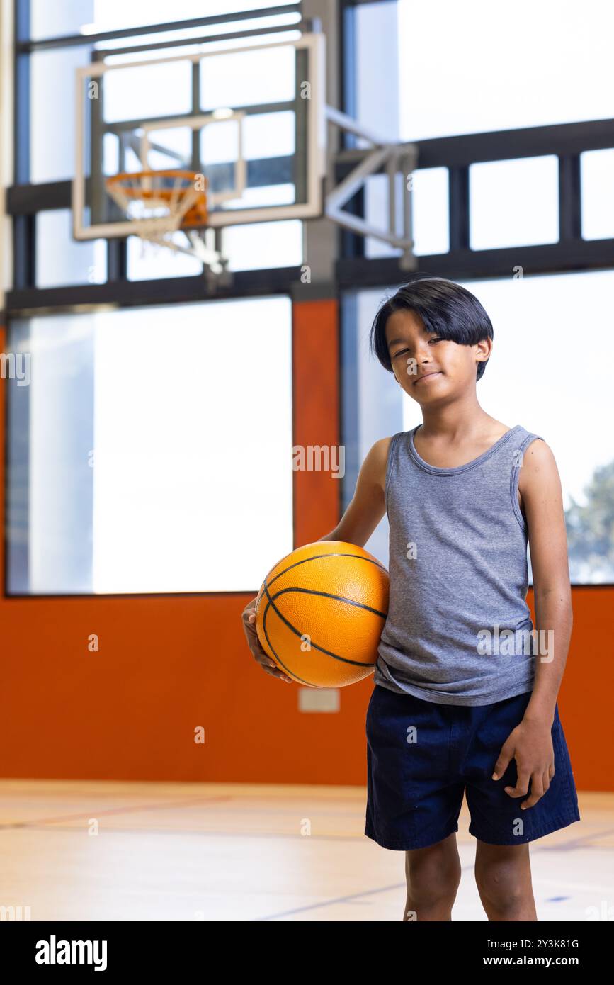 In school gym, boy holding basketball and standing near hoop, looking ...