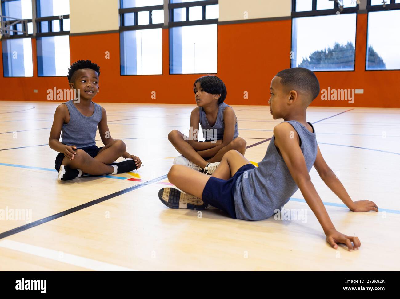 In school gym, three multiracial boys sitting on floor, wearing sports ...