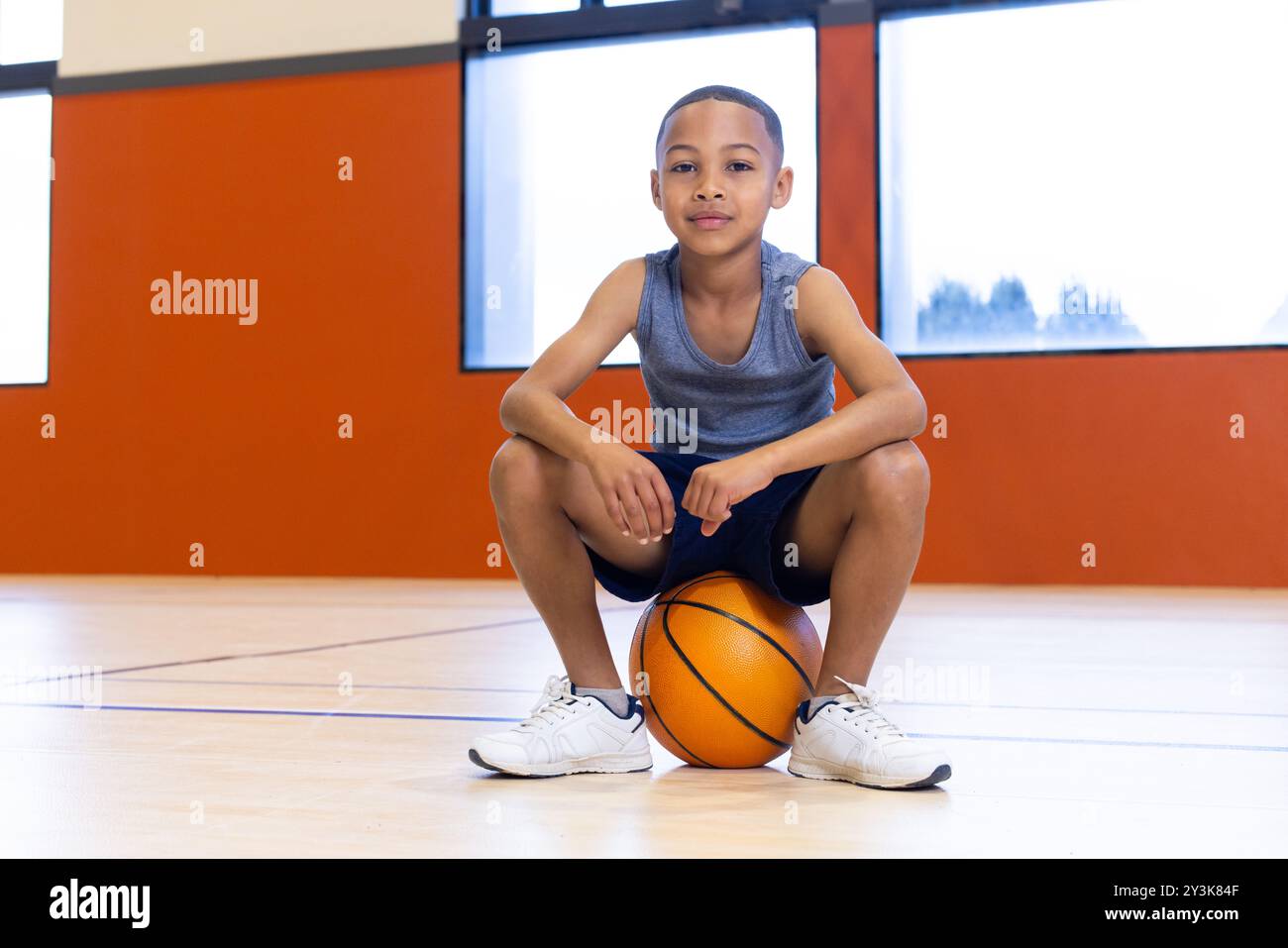 In school gym, boy sitting on basketball, looking confident and relaxed ...
