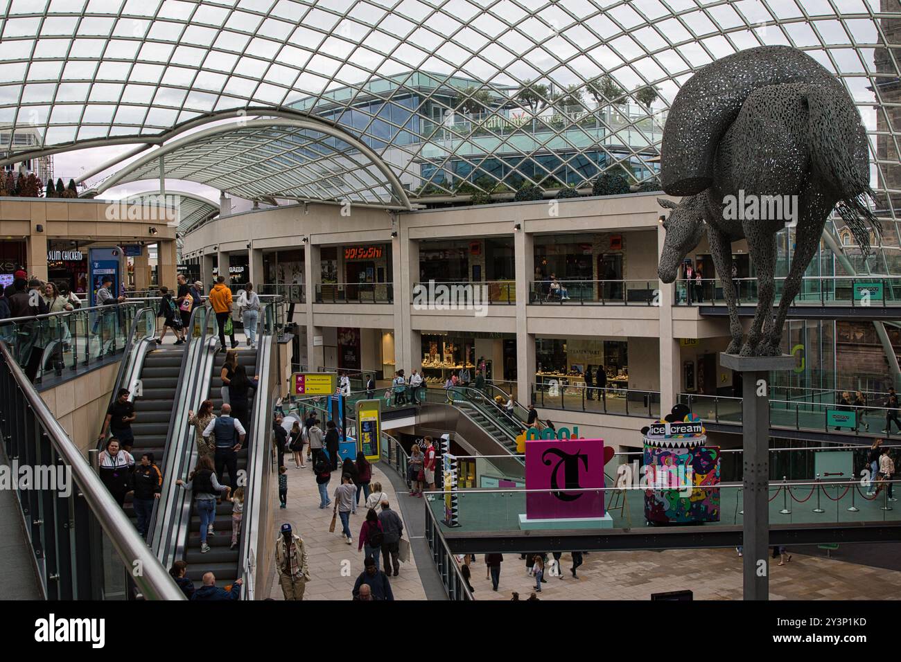 A bustling shopping mall with a glass roof, featuring escalators and a ...