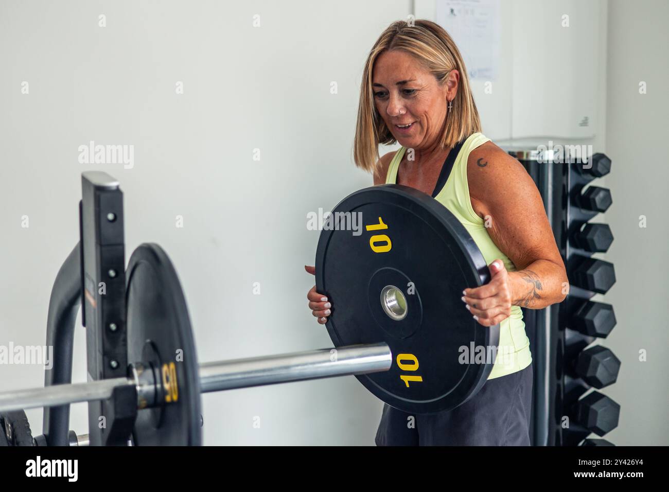 smiling mature blonde tattooed woman is placing a weight plate on the ...