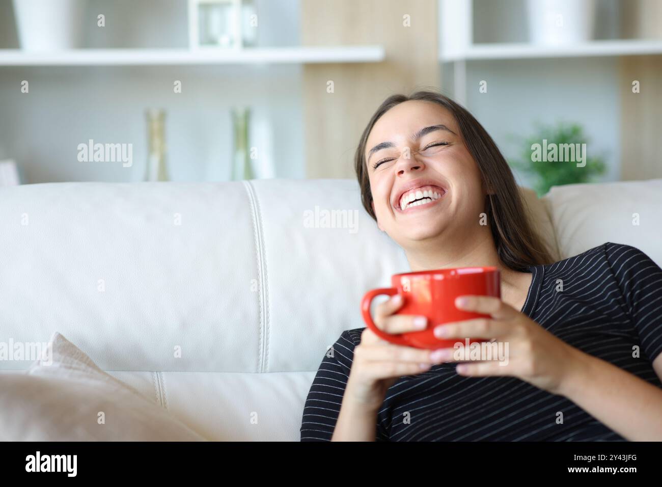 Funny woman laughing drinking coffee alone sitting on a couch at home ...