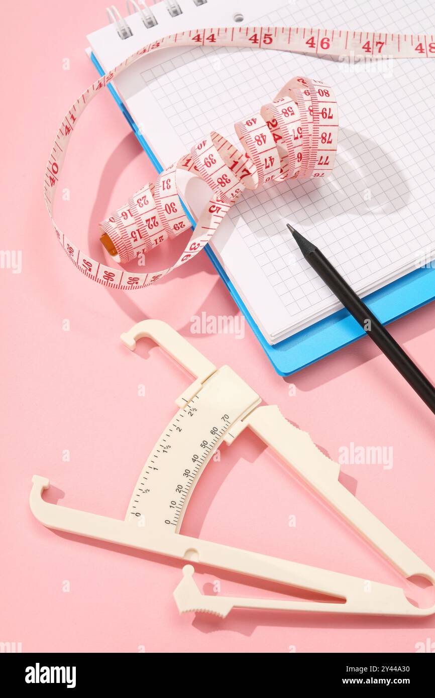 Body fat caliper, measuring tape, notebook and pencil on pink ...