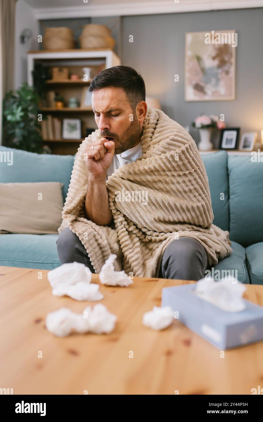 Man dealing with flu symptoms, coughing and staying warm on the couch ...