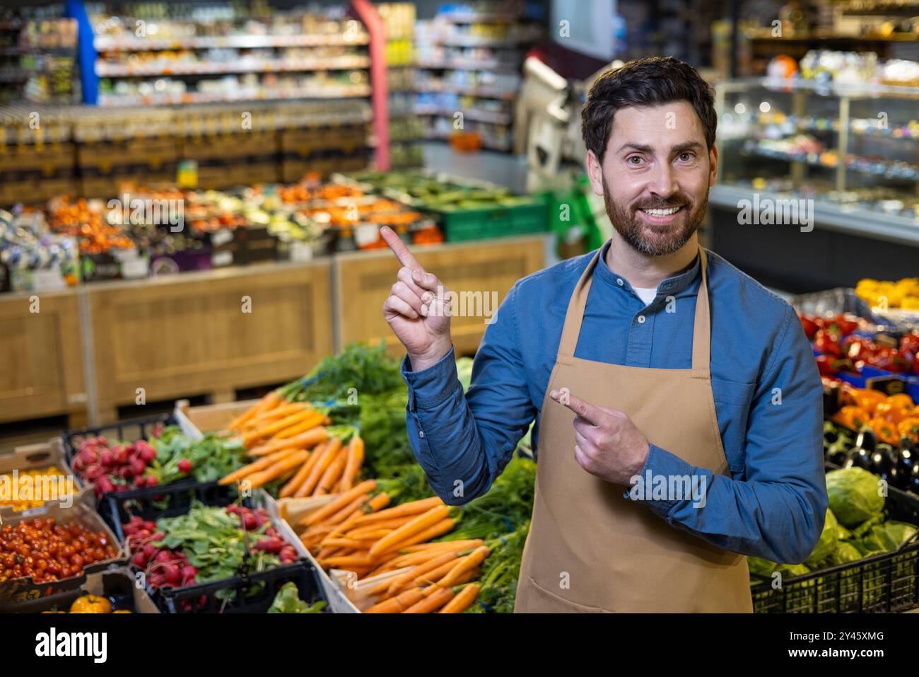 Smiling grocery store employee in apron pointing proudly at colorful ...