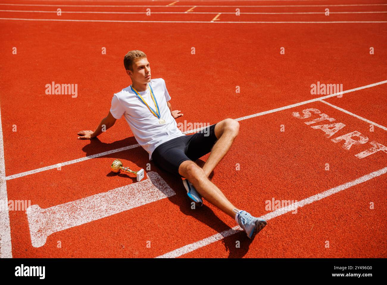 A young male athlete sitting on a running track, smiling and relaxed ...