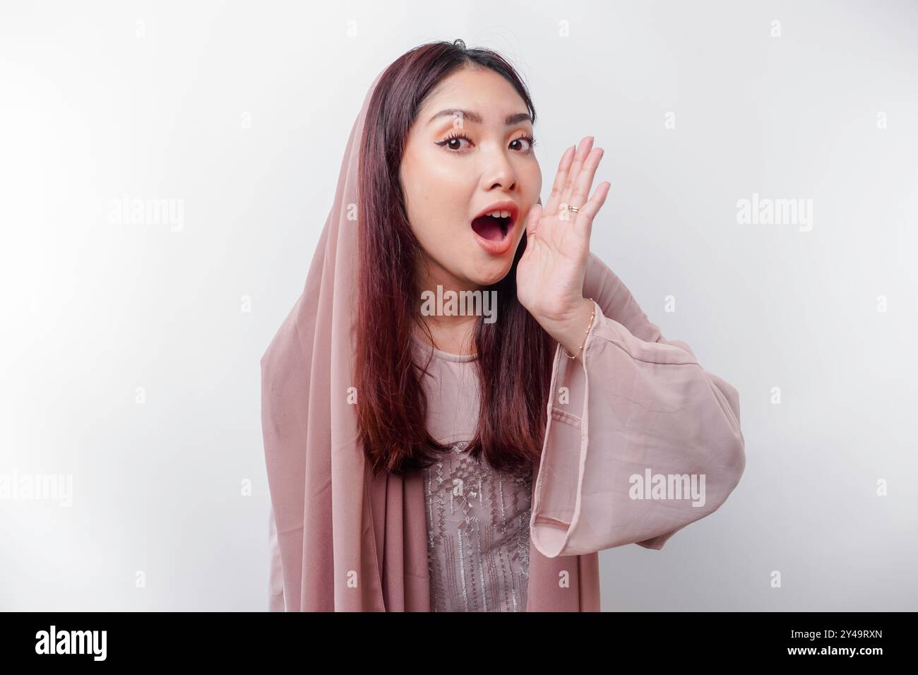 Young beautiful Asian Muslim woman wearing a headscarf shouting and screaming loud with a hand ...