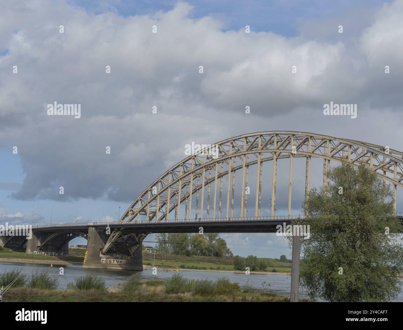Large steel bridge over a river, trees in the foreground and clouds in ...