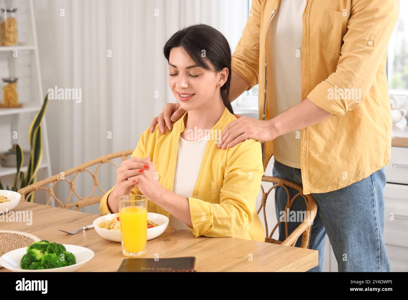 Religious young woman with her husband praying before dinner in kitchen ...