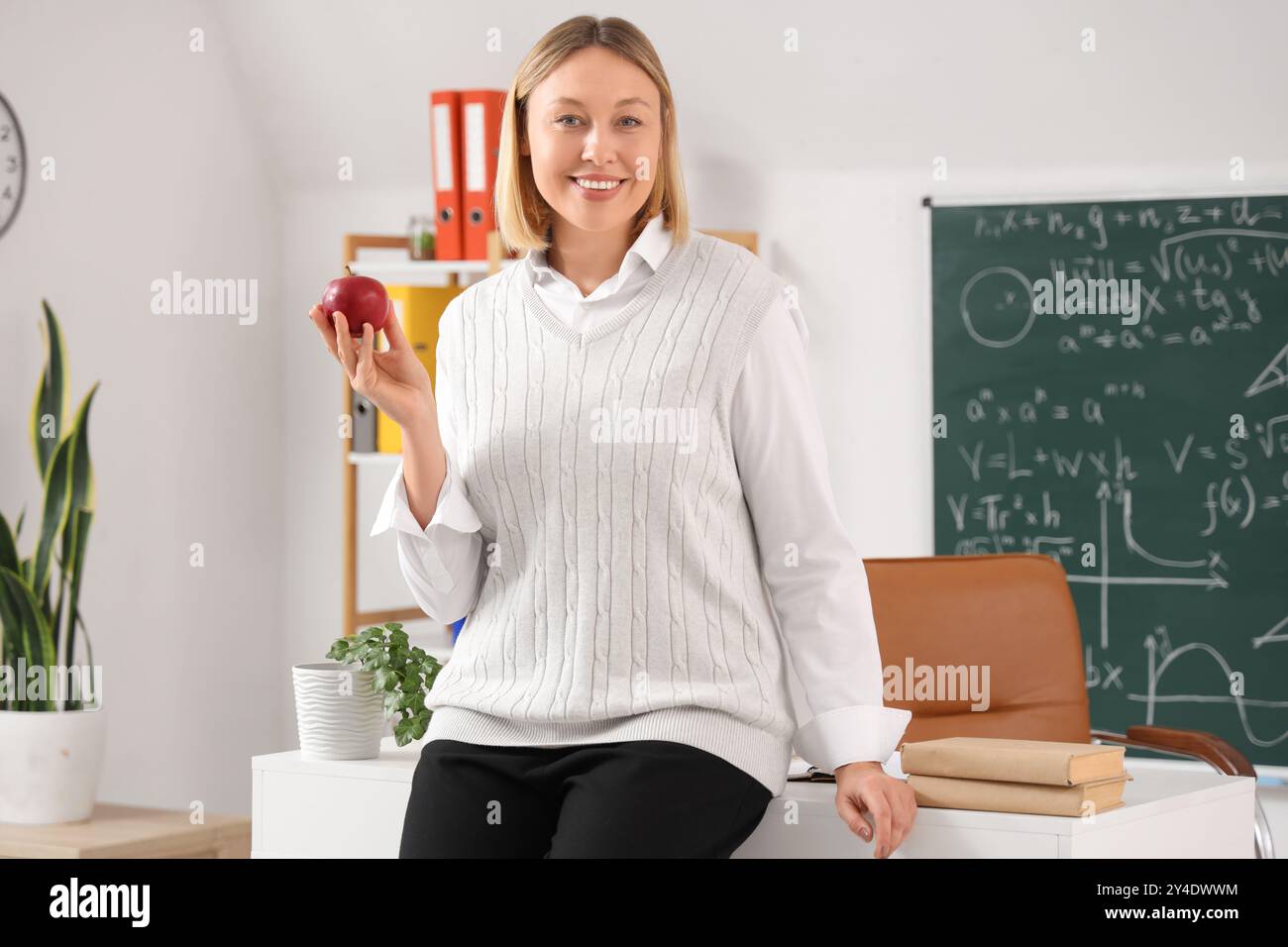 Mature female math teacher holding apple near table with books in ...