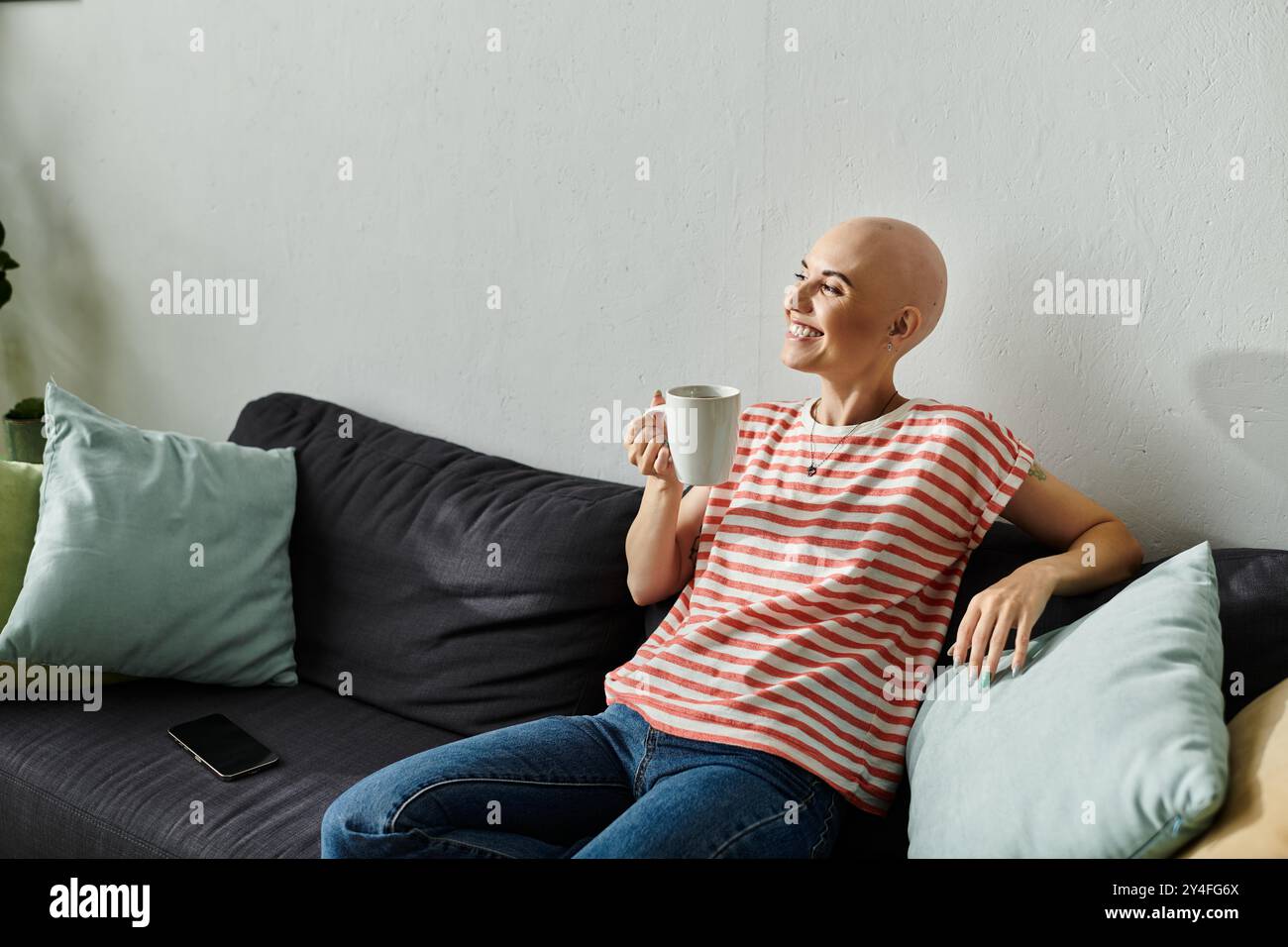 A bald woman smiles while sipping tea, relaxed on a comfortable couch ...