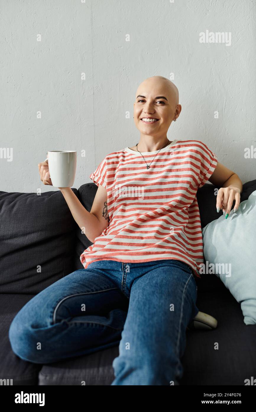 A cheerful bald woman with alopecia sits on a couch, holding a mug and ...