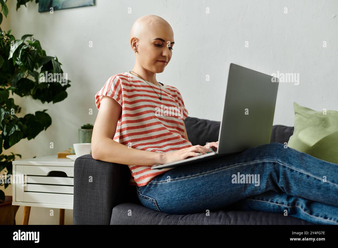 A young bald woman sits comfortably on a couch, working on her laptop ...