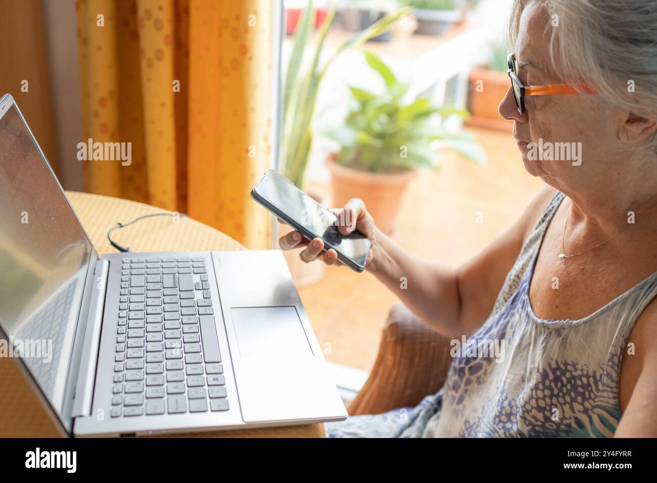 A senior woman multitasks by using a smartphone and a laptop while ...