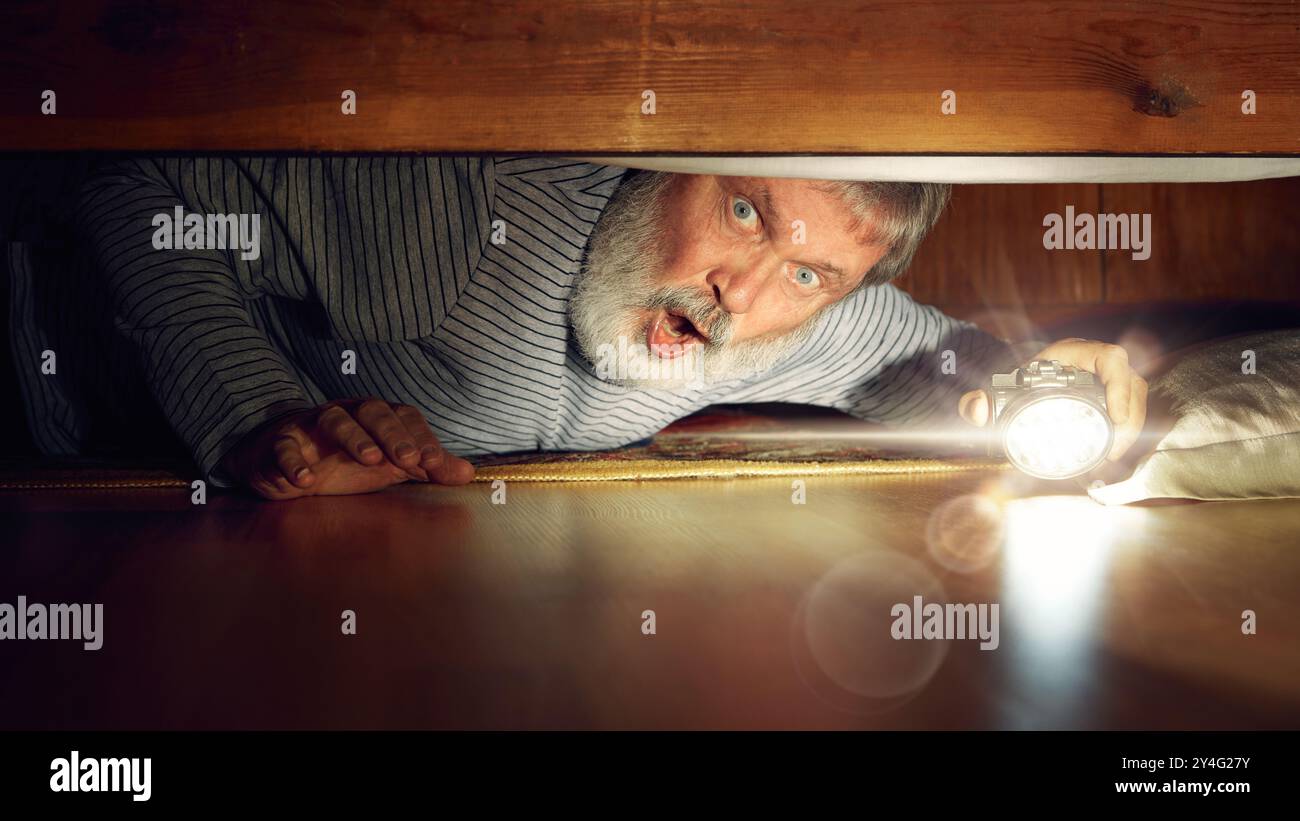 Emotional senior man looking under bed with flashlight, expressing ...
