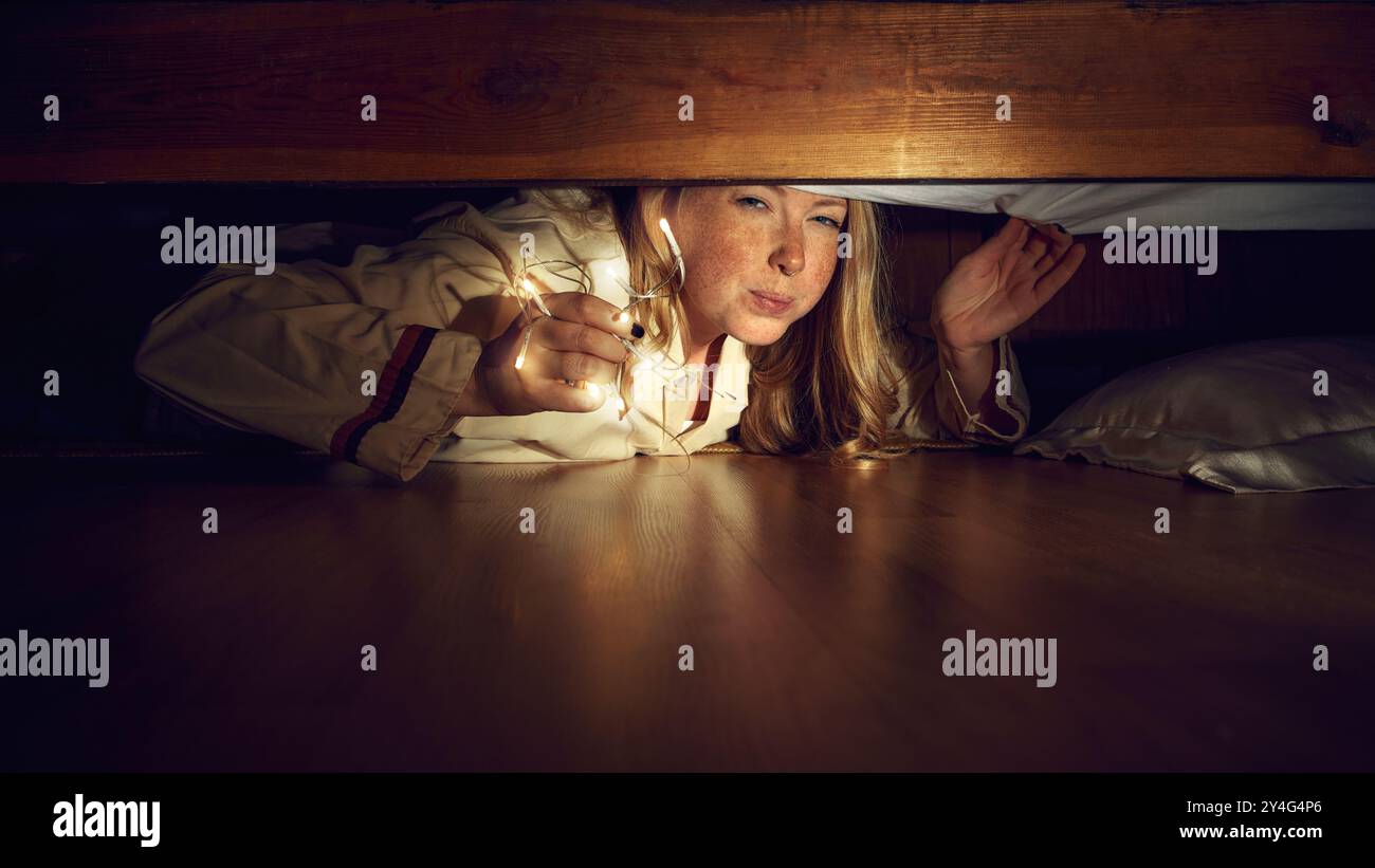 Young emotive woman lying on floor and looking under bed with light ...