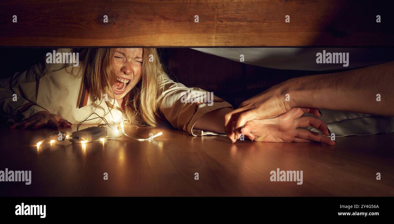 Young fearful woman lying under bed with light while male hand grabbing ...