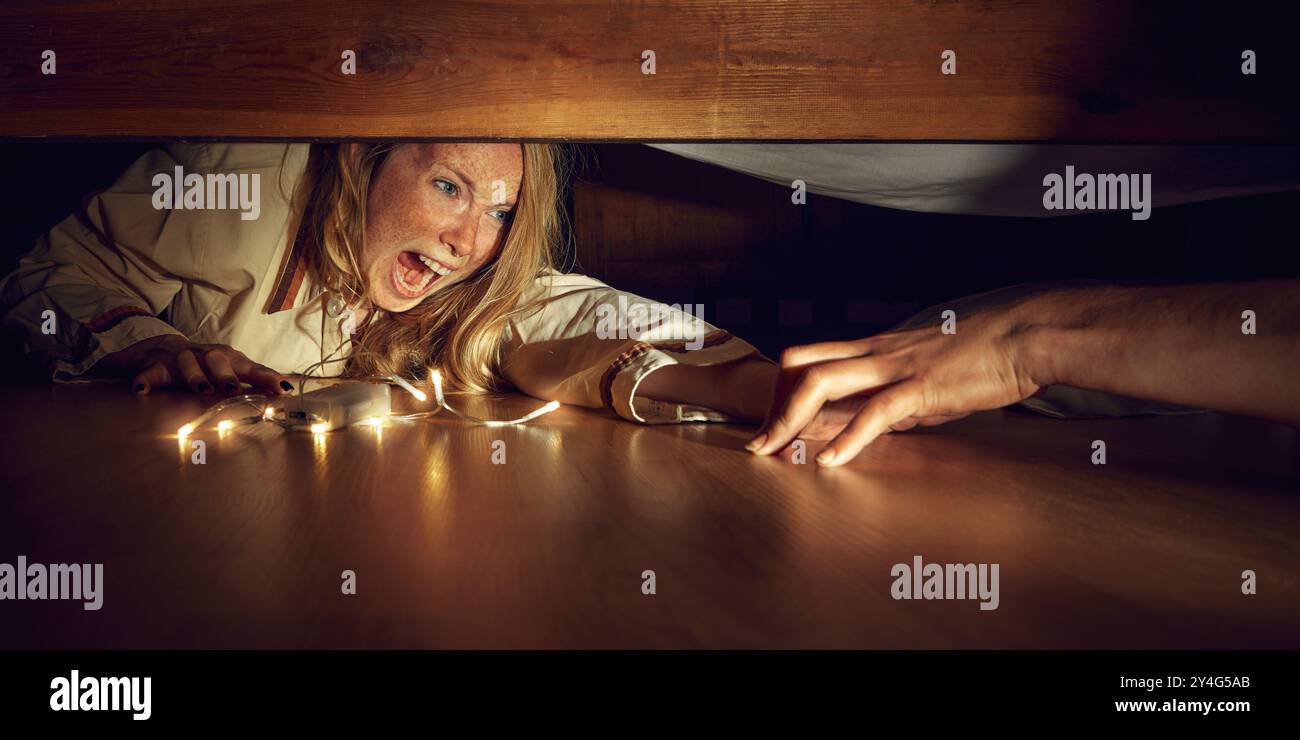 Young fearful woman lying under bed with light while male hand grabbing ...