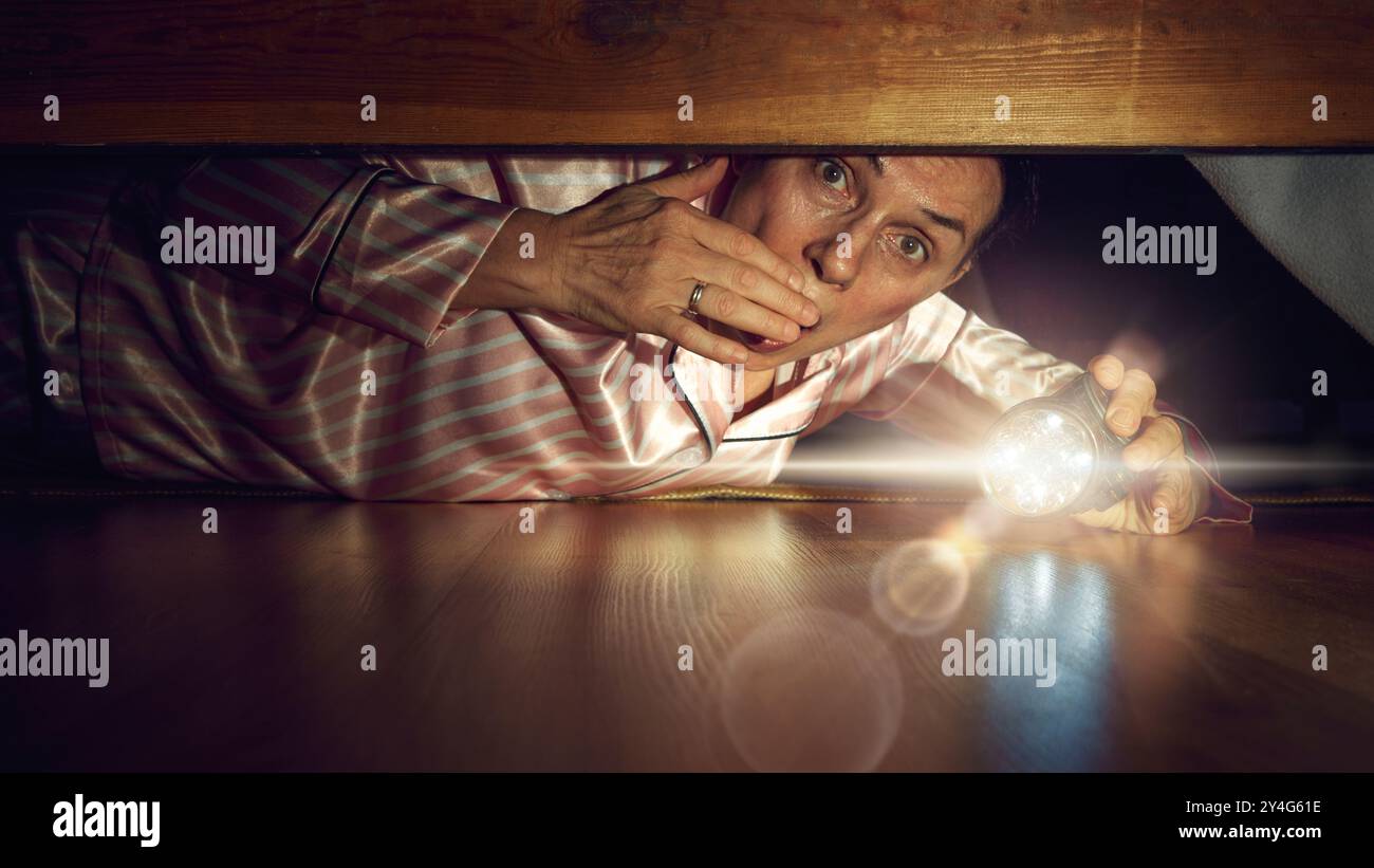Emotional senior woman looking under bed with flashlight, expressing ...