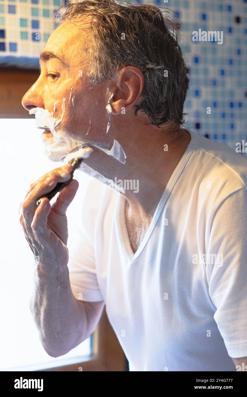 A middle-aged man shaves with a razor in a bathroom. He is ...