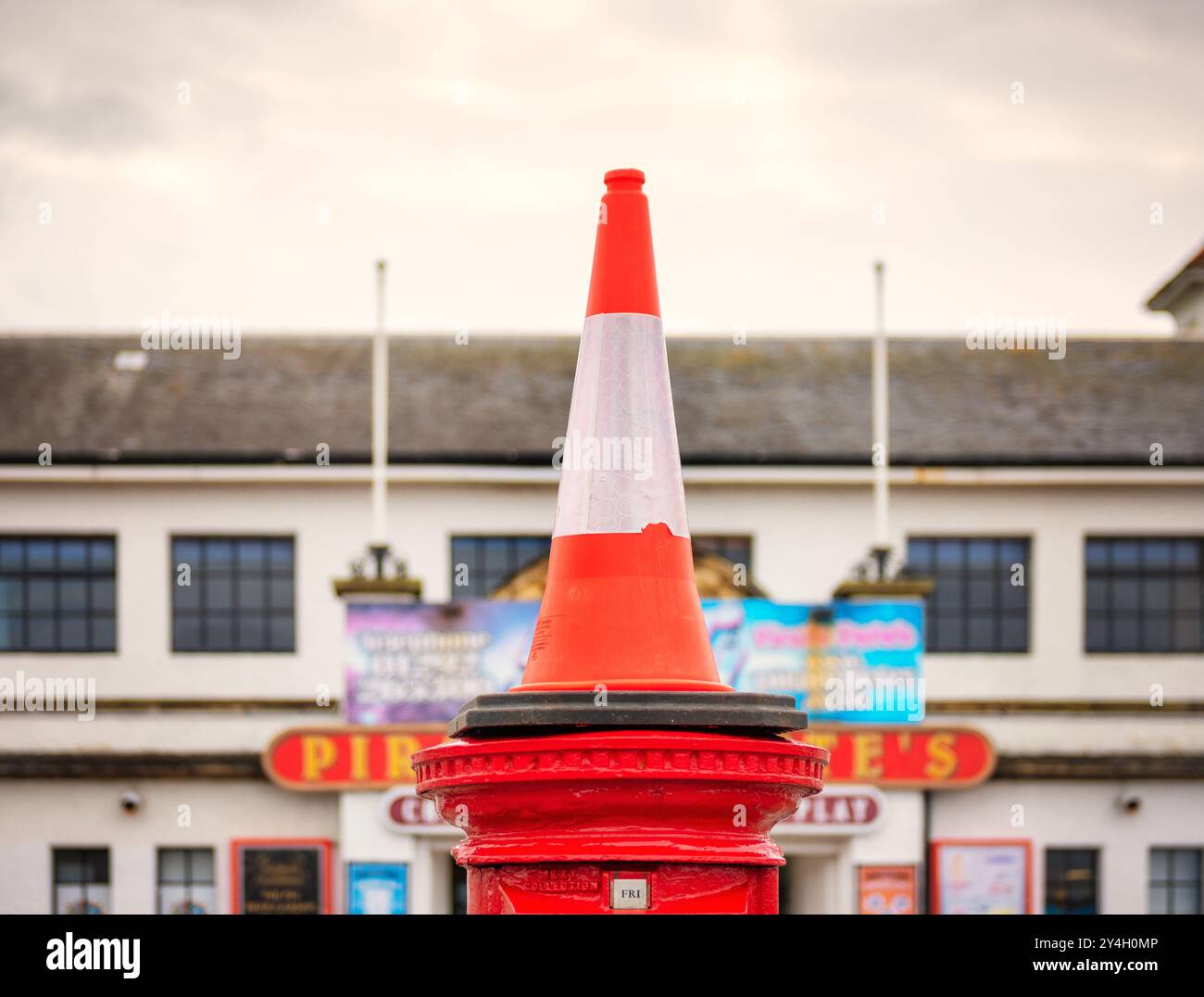 An orange traffic cone on top of a red post box in front of a run down ...