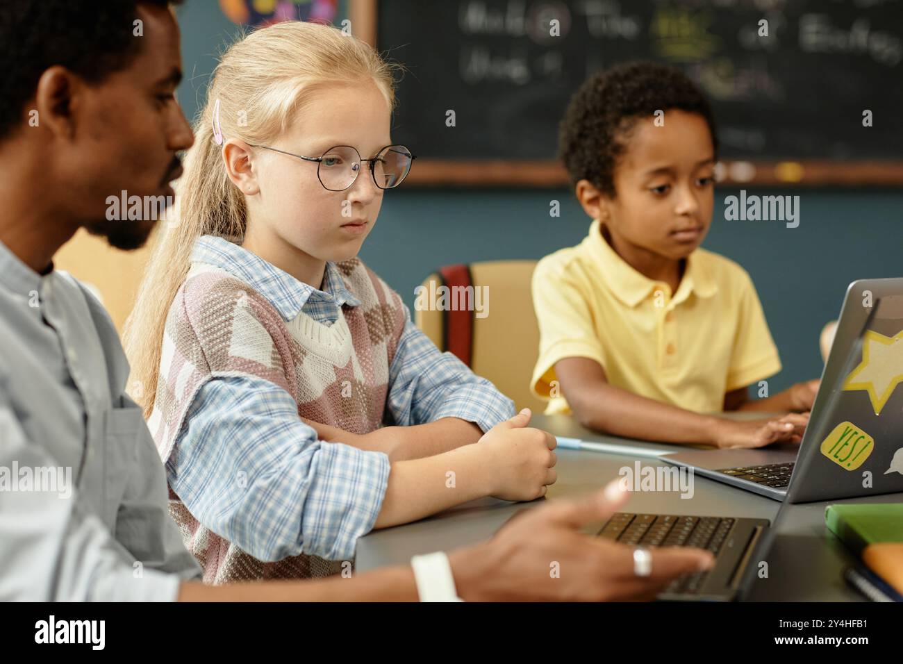 Side view of young blonde girl sitting at desk with laptop listening ...