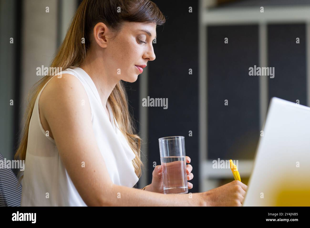 Holding glass of water, woman writing notes while working on laptop ...