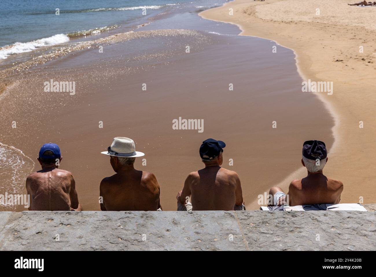 Four elderly men sit on a ledge overlooking the beach in Cascais ...