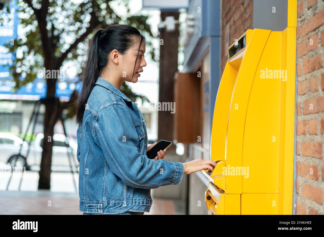 A side view of a beautiful Asian woman in a denim jacket inserting her debit card into an ATM to ...