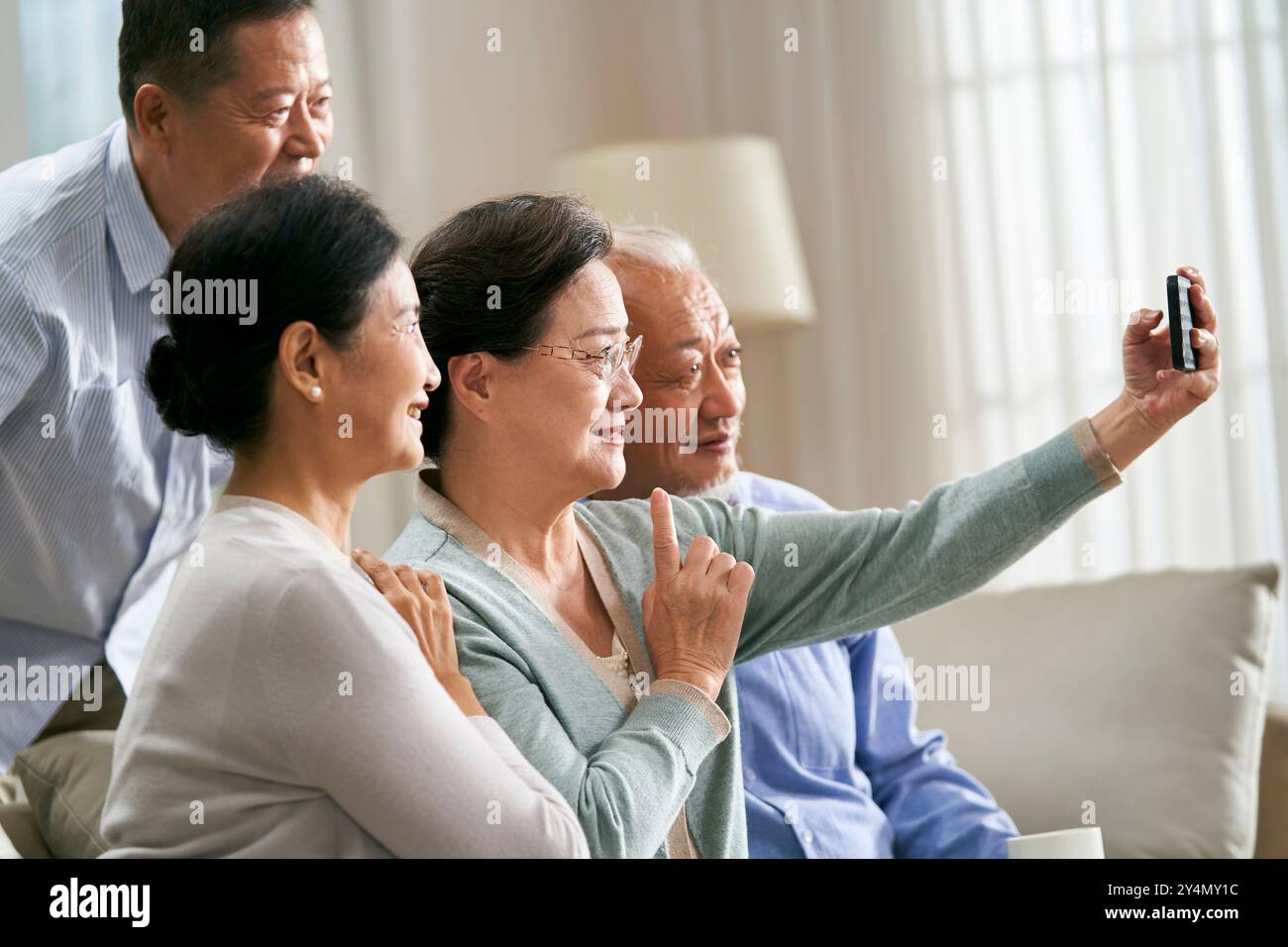 group of happy senior asian people two couples sitting on family couch at home taking a selfie ...