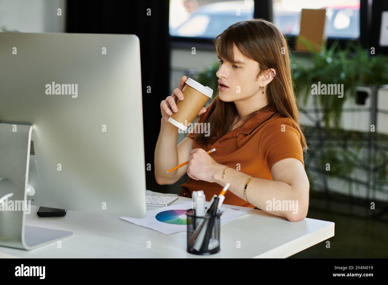 Focused non binary individual holds coffee while working at a desk amid ...