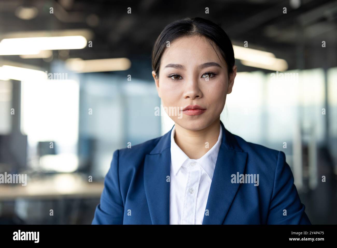 Confident businesswoman wearing blue suit looking directly at camera in modern office setting ...