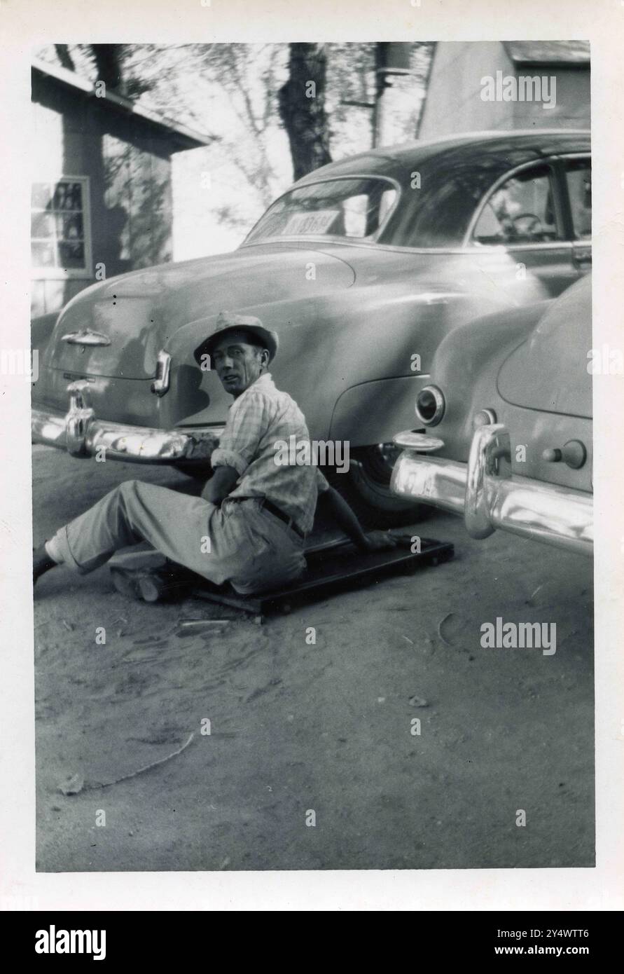 A mechanic works on the underside of a 1940s car, using a creeper to ...