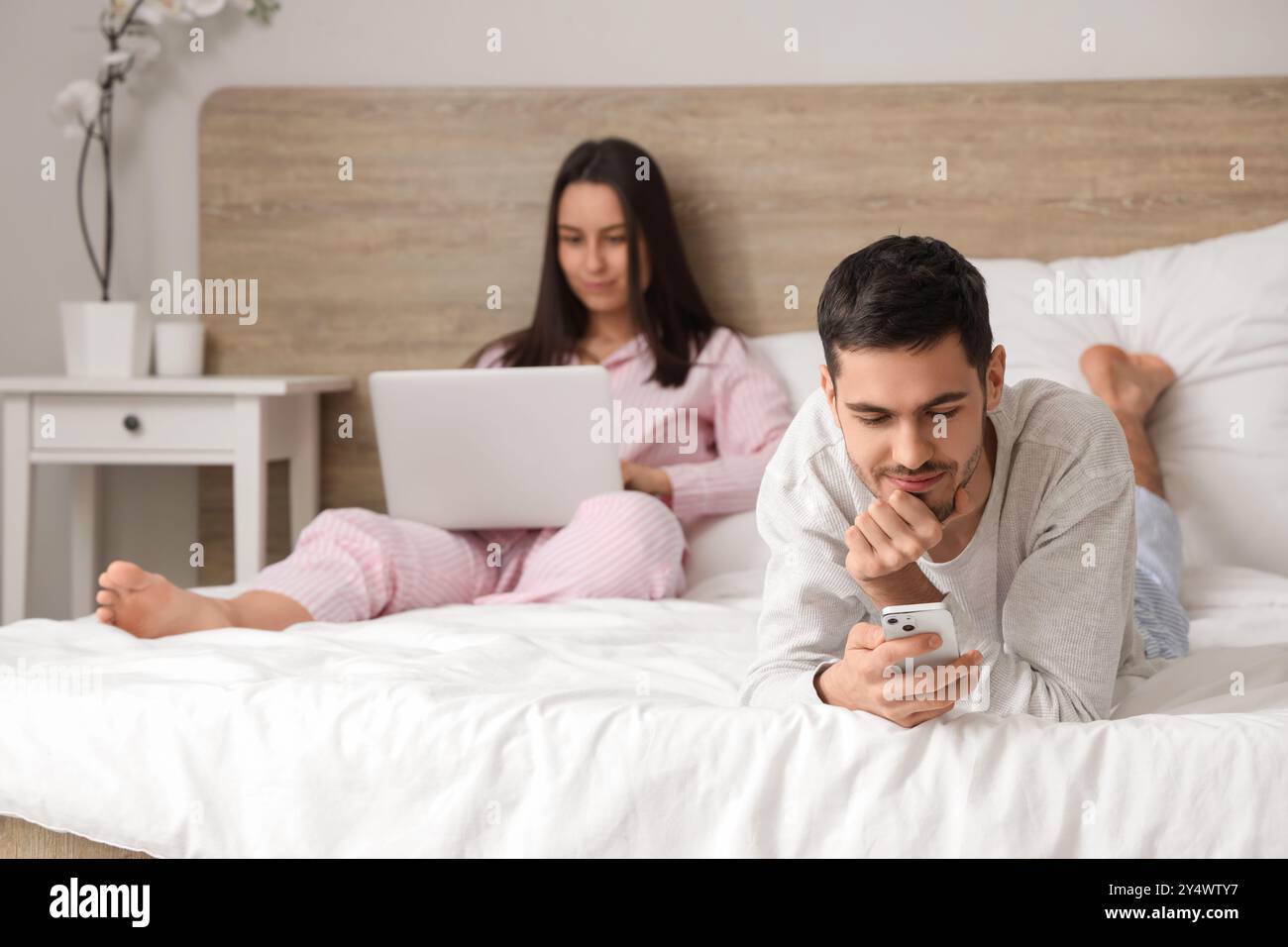 Young guy with mobile phone and his girlfriend with laptop lying on bed ...