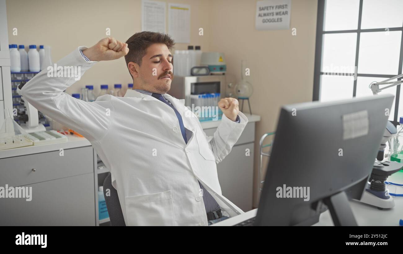 A young man in a lab coat stretches at his desk in a well-equipped ...
