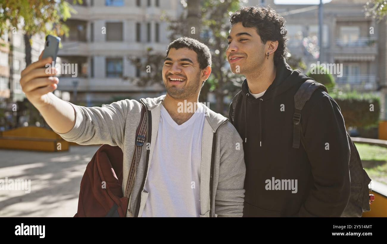 Two smiling men taking a selfie together in a sunlit urban park setting ...