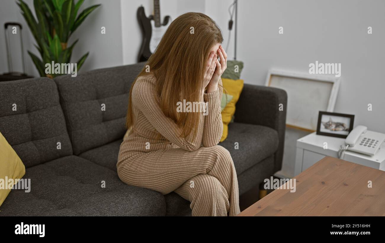 A distraught young woman sitting in a modern living room, covering her ...