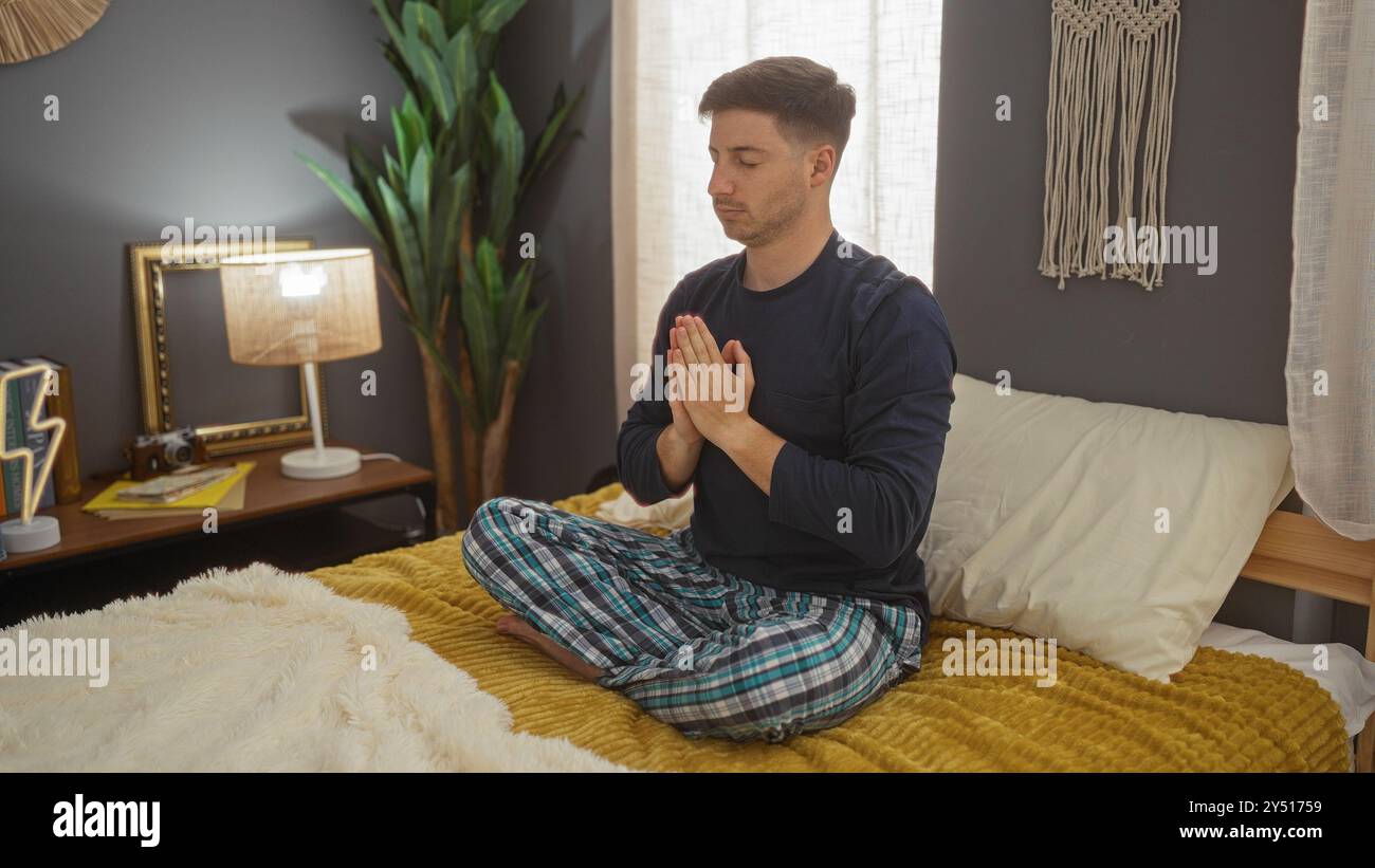A young hispanic man sits in meditation with hands in prayer position ...